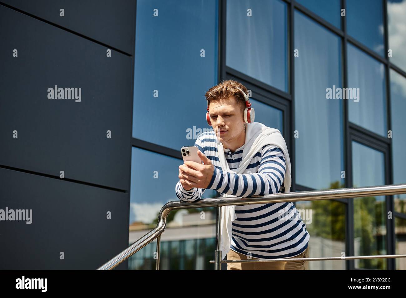 A stylish young man leans on a railing, using his phone and listening ...