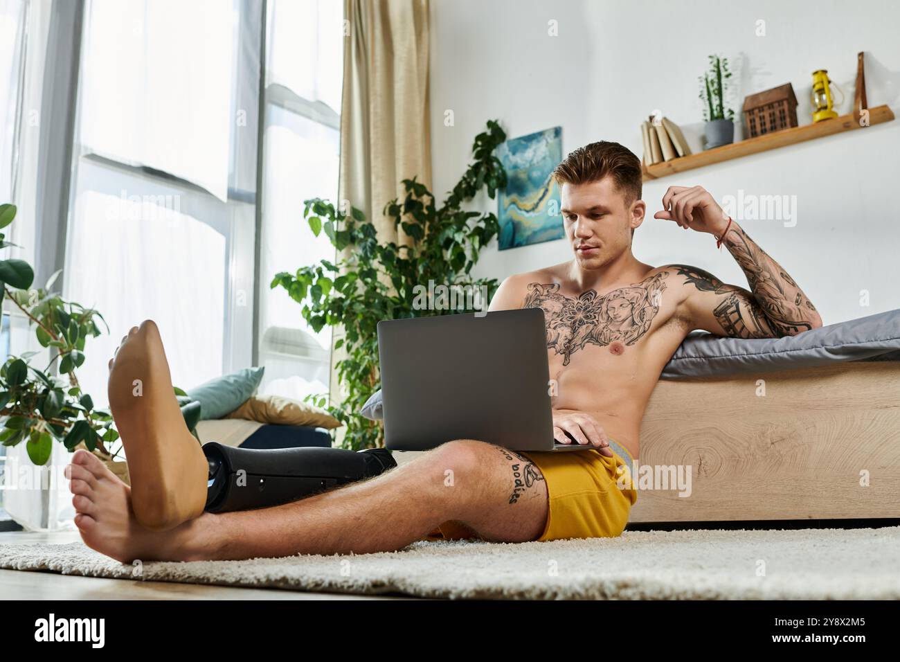 A young man with tattoos sits on a rug, focused on his laptop while ...