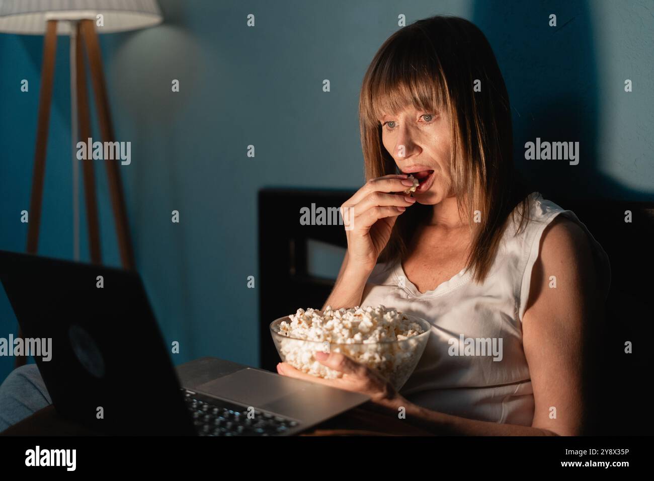 Mature cute woman eating popcorn while sitting on bed under blanket and ...