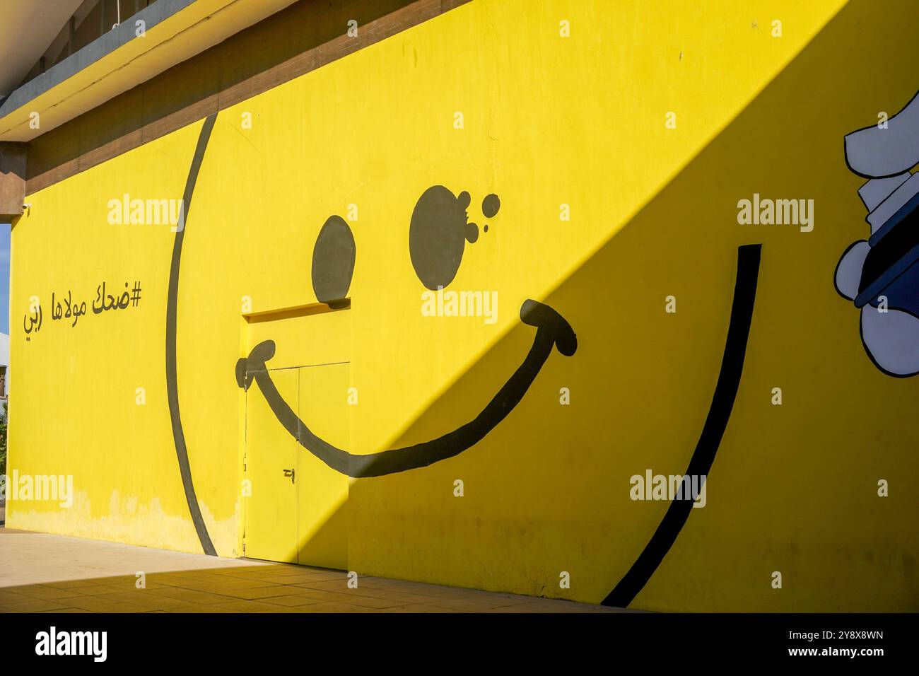 A vibrant yellow wall adorned with an oversized smiley face mural casts ...