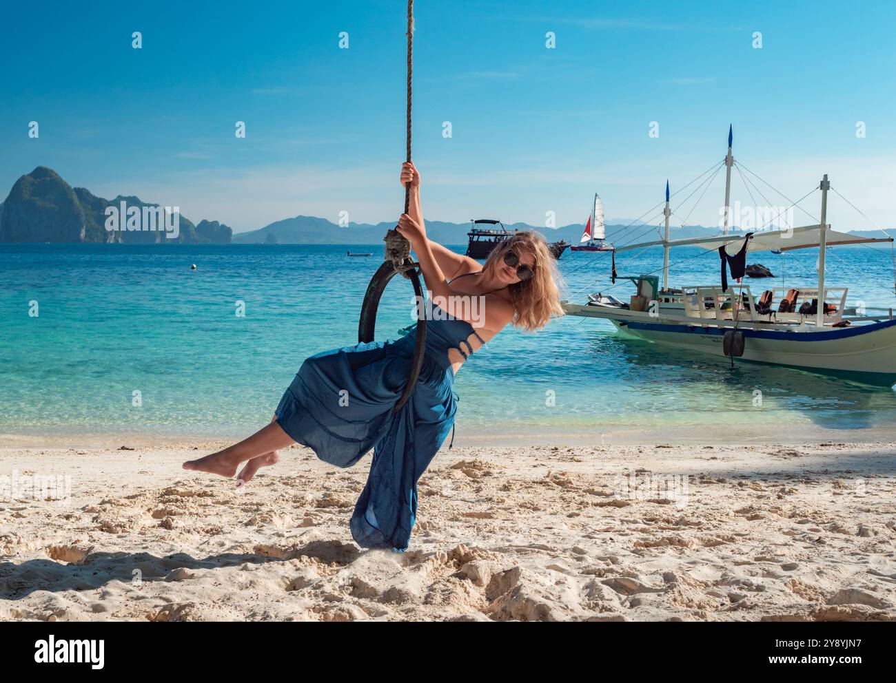 A woman with long blonde hair, wearing a blue dress, swings on a tire ...