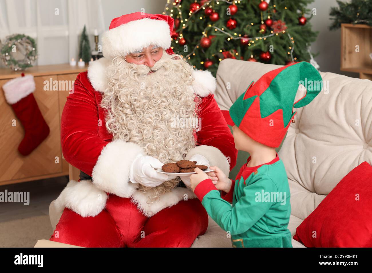 Santa Claus and cute little elf eating cookies at home on Christmas eve ...