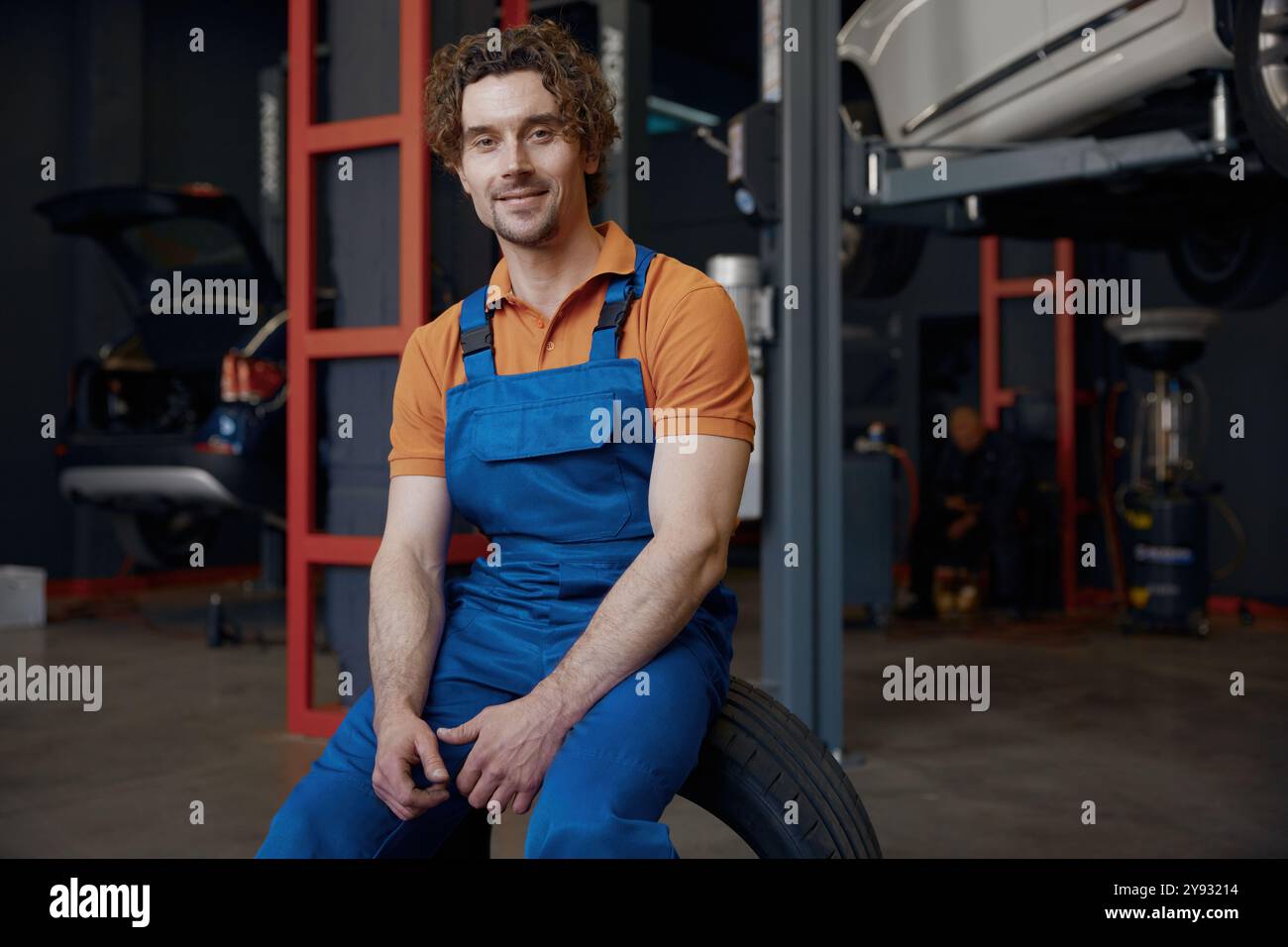 Happy smiling relaxed male mechanic sitting on tire in garage car ...