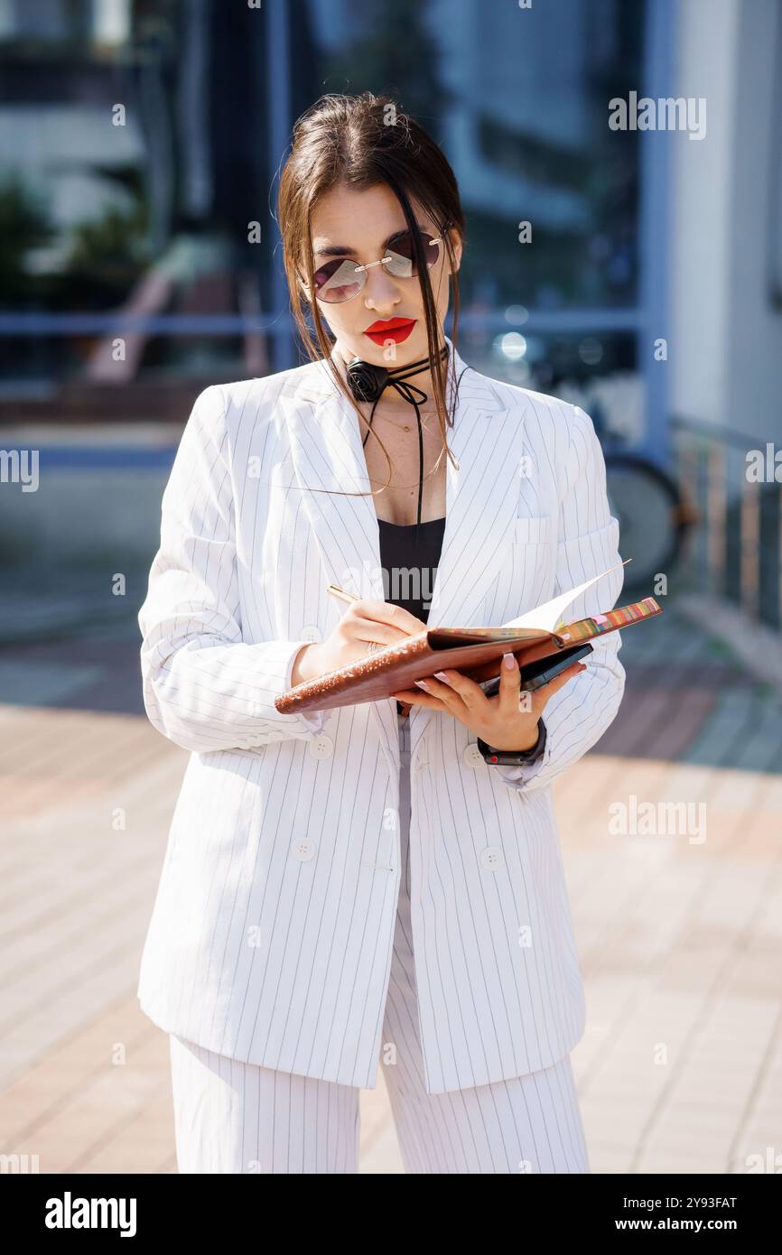 Stylish businesswoman jotting down notes in a notebook outdoors with ...