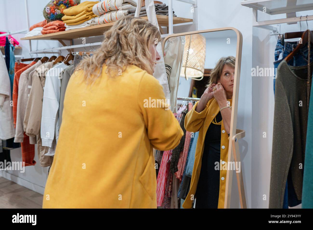 Two women are in a clothing store, one trying on a yellow coat ...
