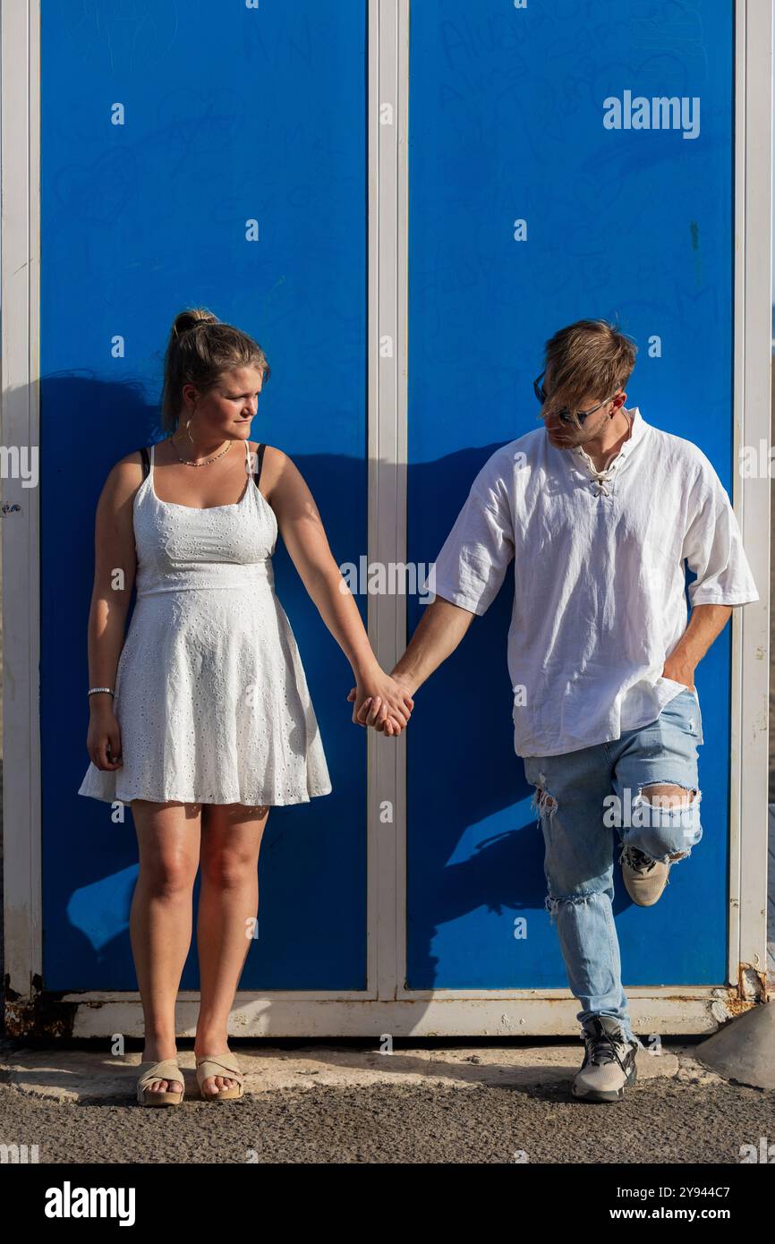 A young couple stands side by side, holding hands in front of a blue ...