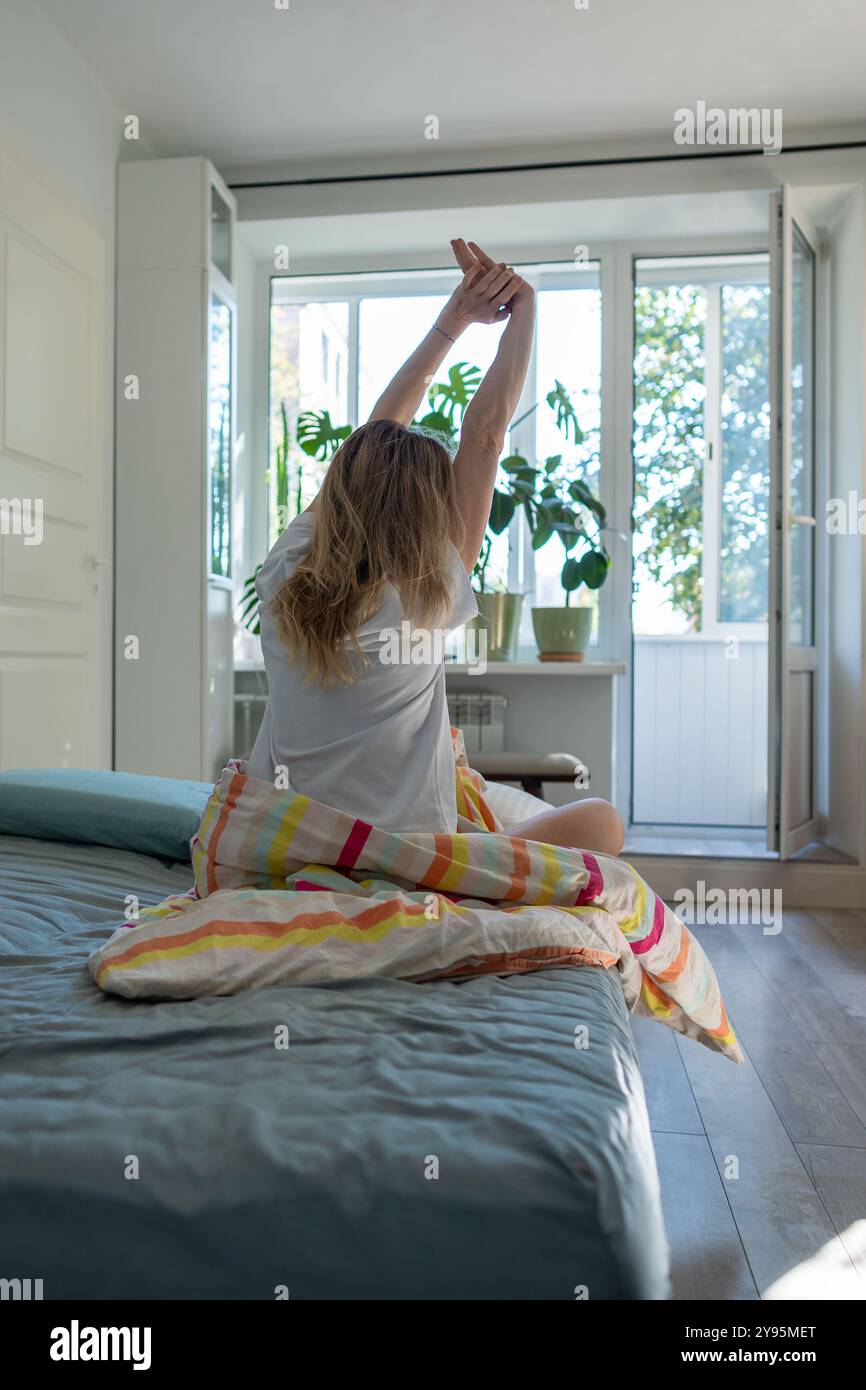Rear view of healthy woman sitting in bed in morning stretching arms ...