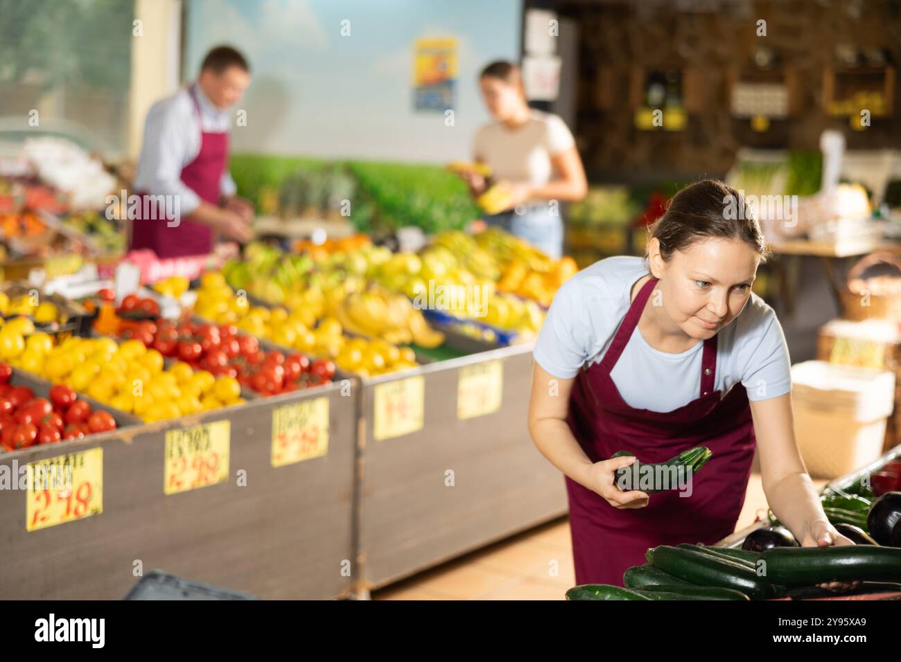 Woman seller working in supermarket and lays out cucumber on counter ...