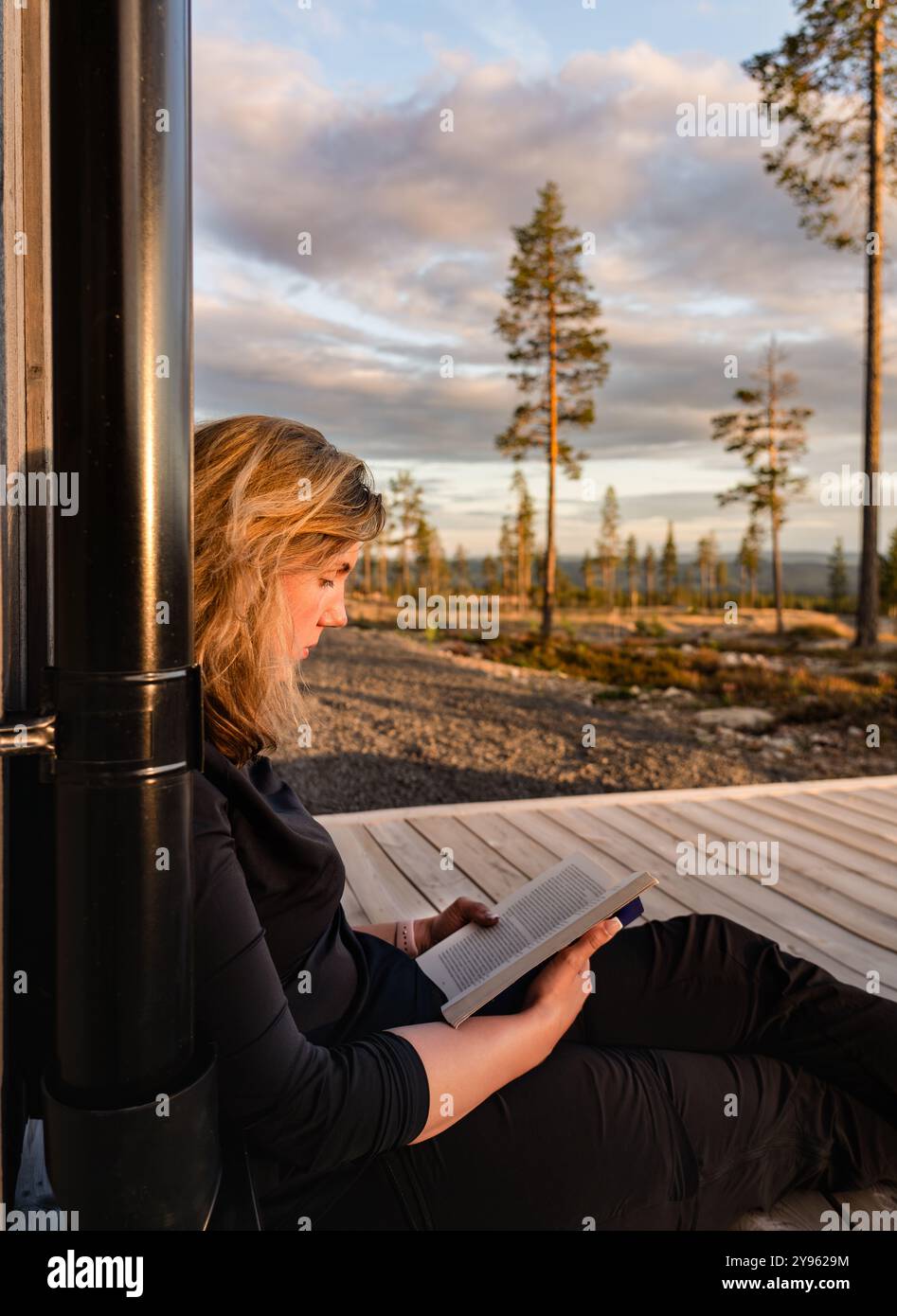 A beautiful woman sits peacefully on a deck, reading a book with ...