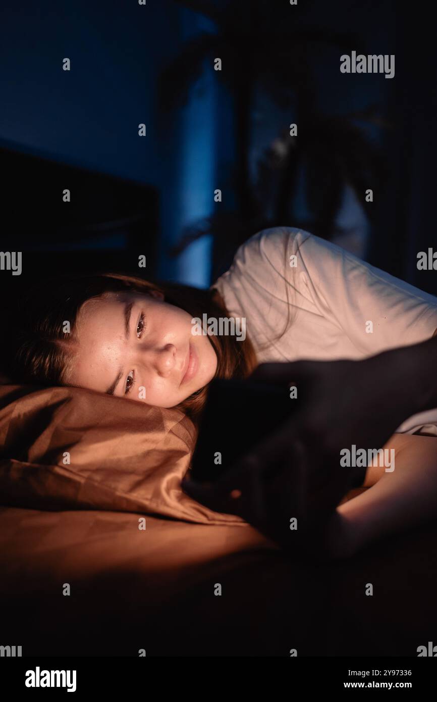 Sleepy teen girl lying in bed using smartphone. Insomnia, addiction concept. Teenage scrolling ...