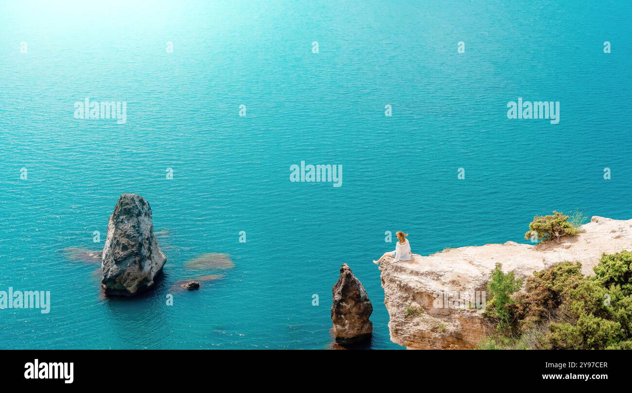 A woman sits on a rock overlooking the ocean. The water is blue and ...