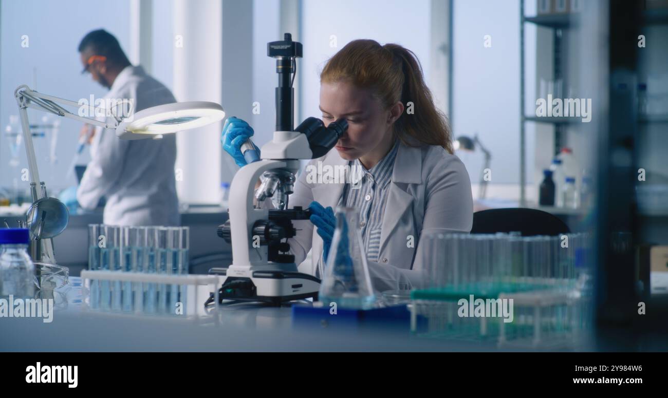 Young female microbiologist looking under microscope, adding new ...