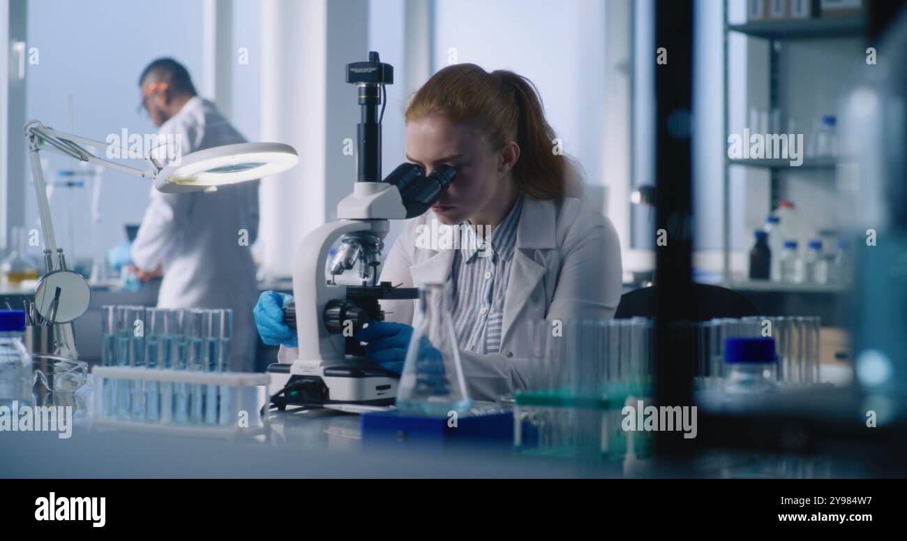 Young female microbiologist looking under microscope, adding new ...