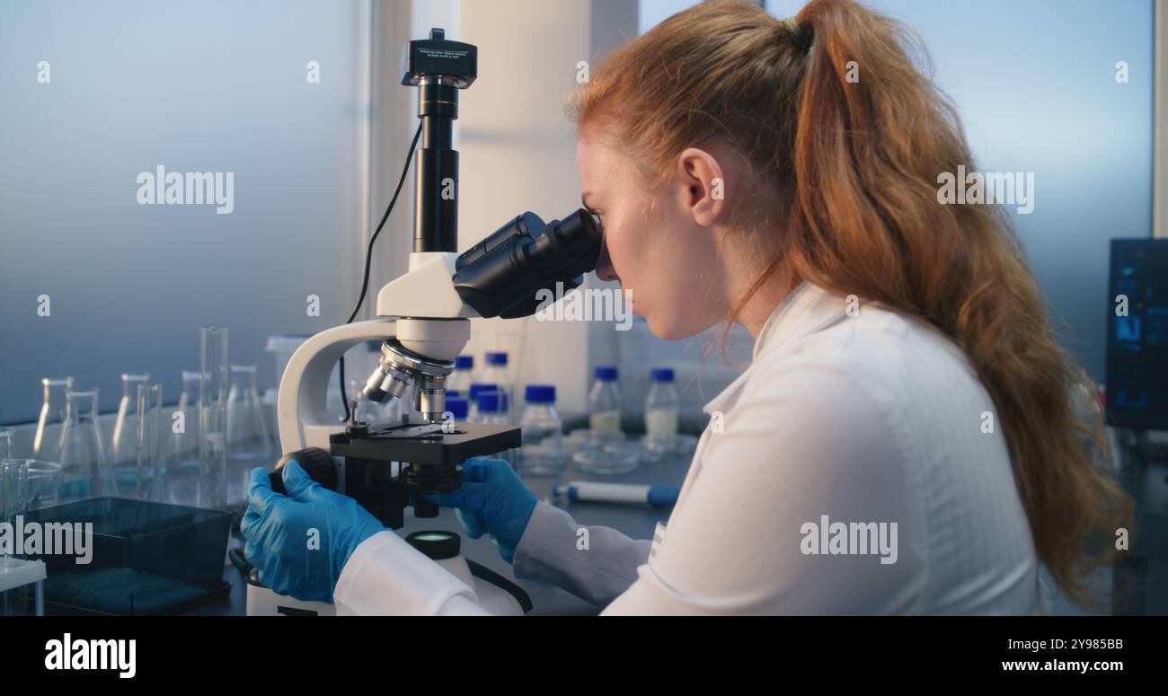 Young female scientist sits in chair, looks under microscope and ...