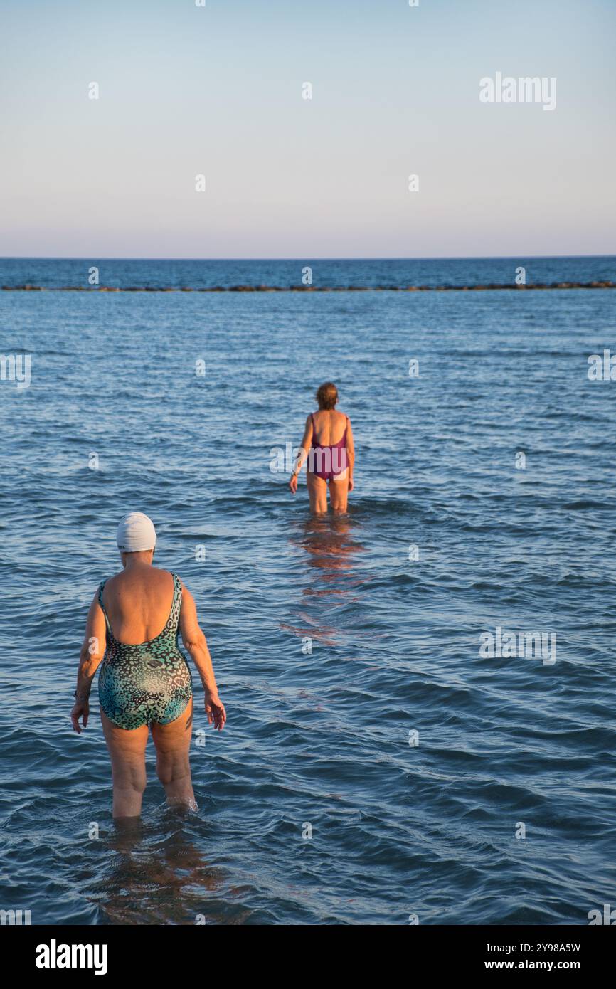 two elderly women entering the waters in Voroklini beach, larnaca just before sunset in Cyprus ...