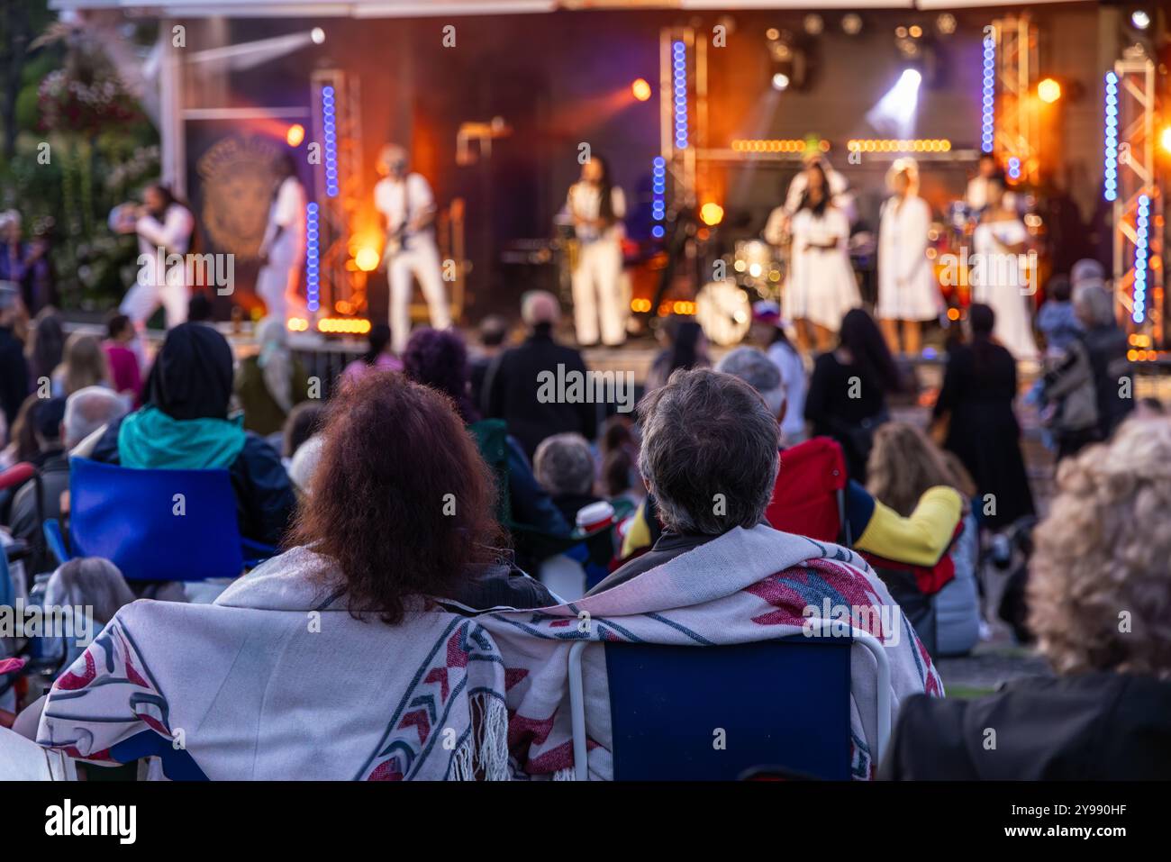 Couple is watching a concert on an outdoor stage at dusk, sitting on ...