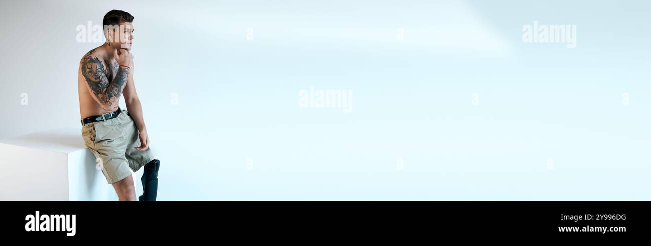 A young man with tattoos thoughtfully poses in a minimalistic studio ...