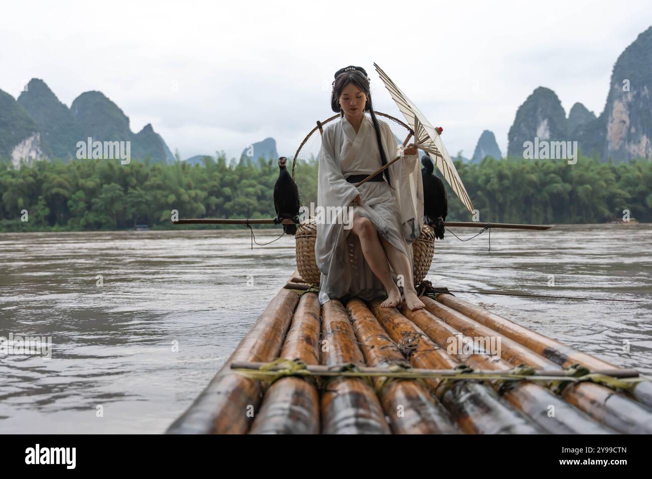 Hanfu girl holding an umbrella poses on bamboo raft on river with two ...