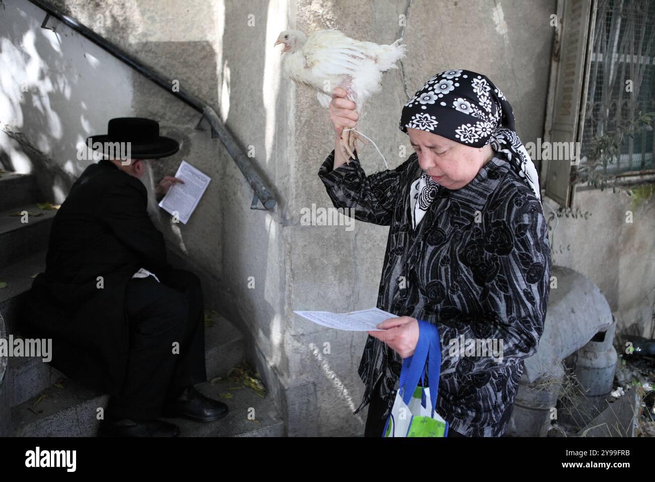 Kapparot, woman swings a rooster over her head, her husband in the back ...