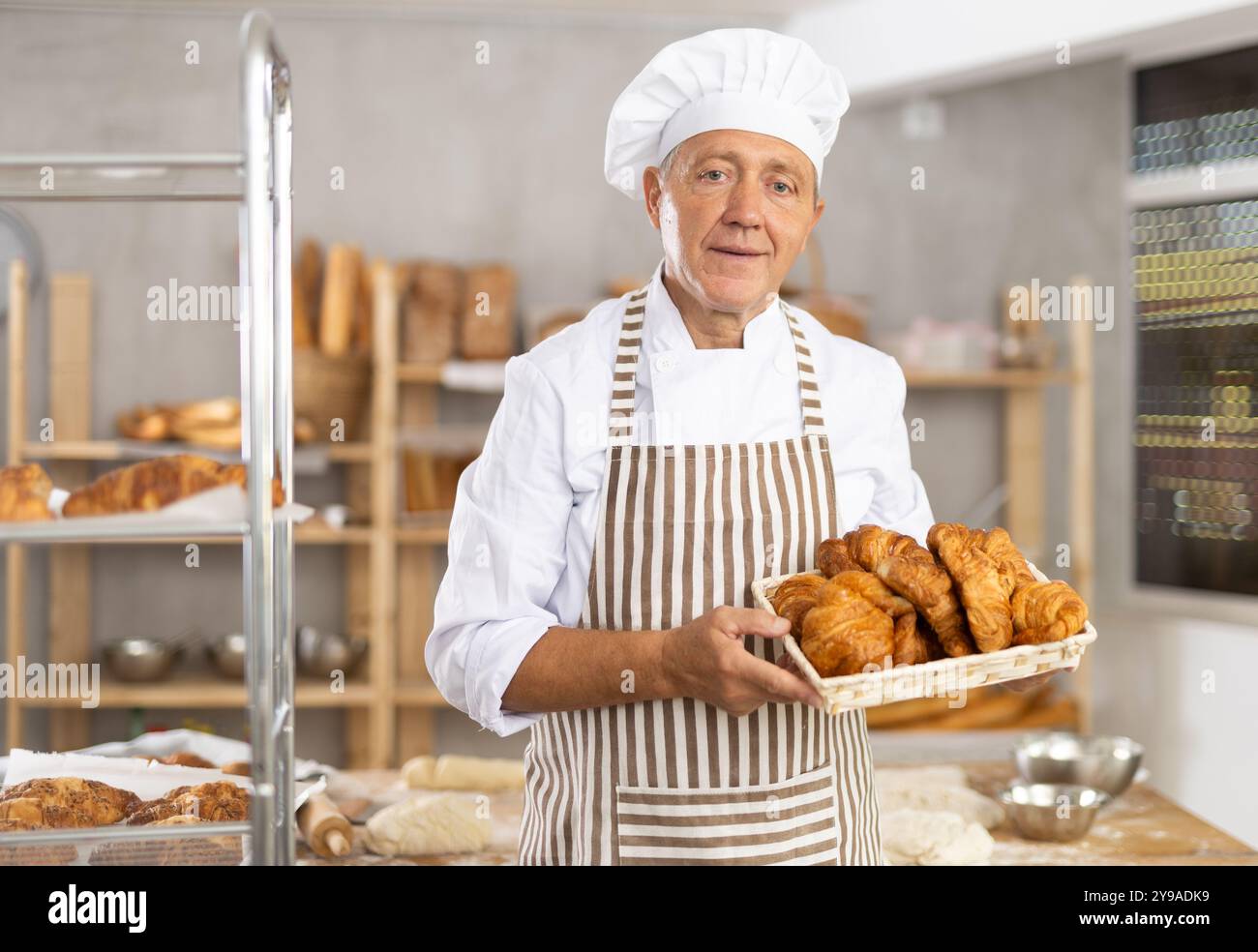 Smiling elderly baker in uniform stands behind counter in small private ...
