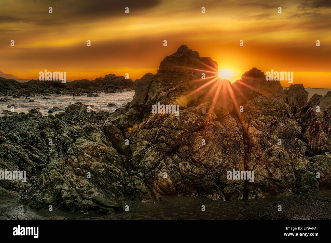 Sun flare poking out behind the rock formation on the coast Stock Photo ...