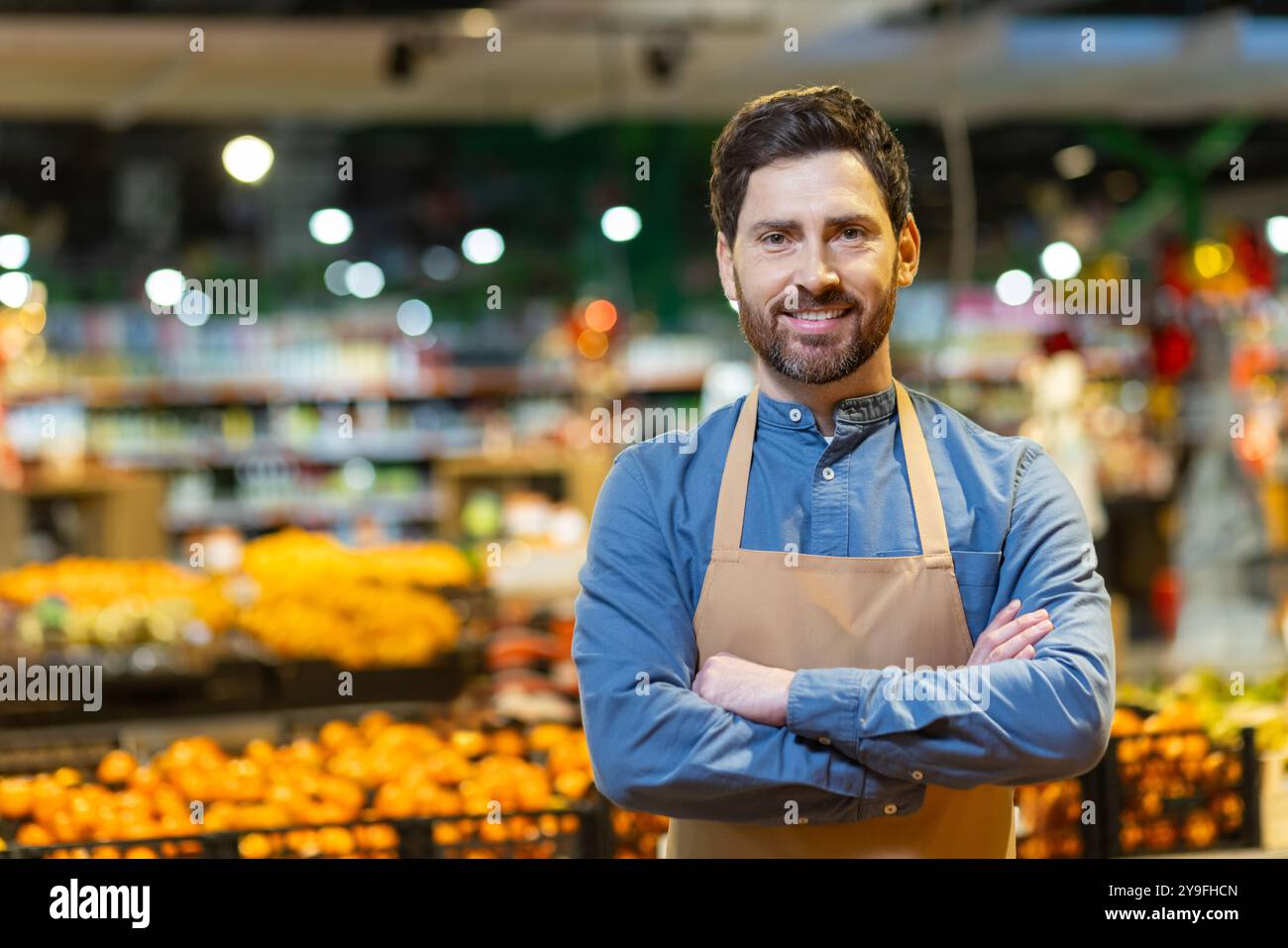 Confident male grocery worker wearing apron stands with crossed arms in ...