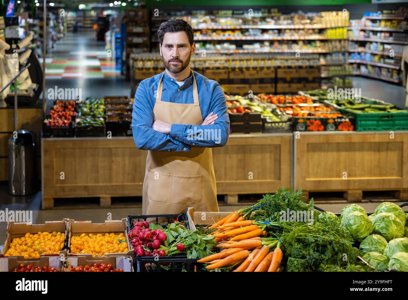 Grocery store worker in apron stands confidently with arms crossed ...