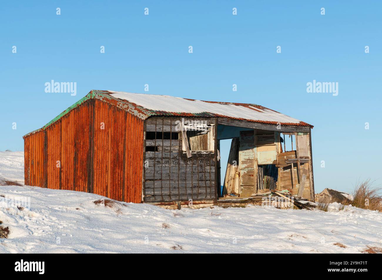 An abandoned, dilapidated shieling sits on a snow-covered landscape, its rusted metal and ...