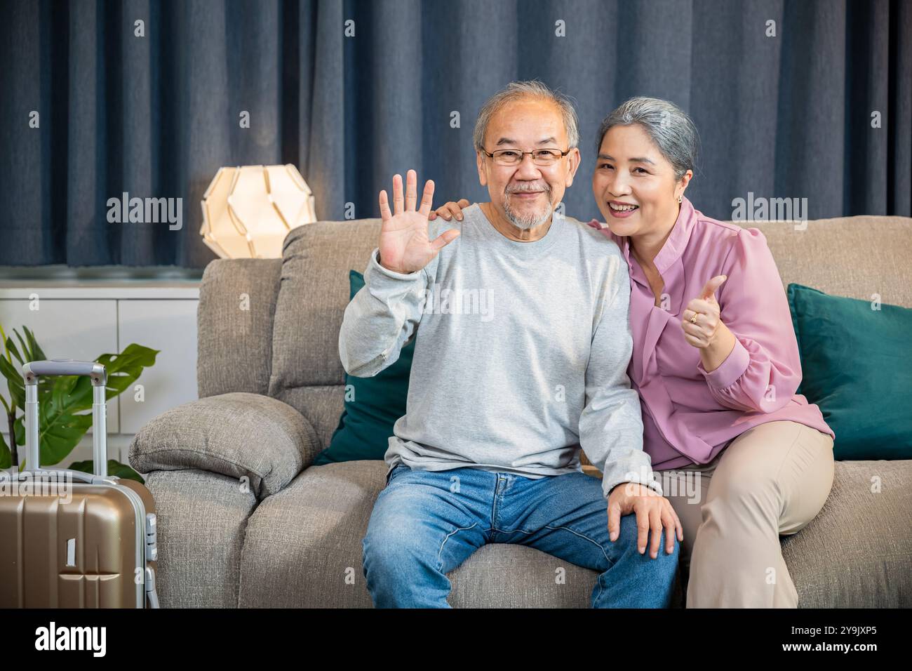 Happy Asian senior couple greeting someone while sitting on a comfortable couch, old man and ...