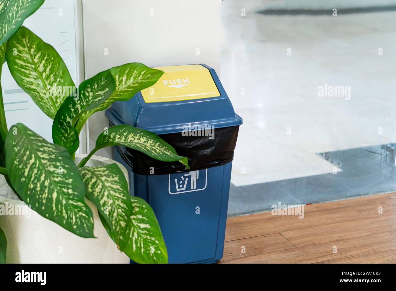 A blue trash bin with a yellow lid labeled PUSH is placed in a corner ...