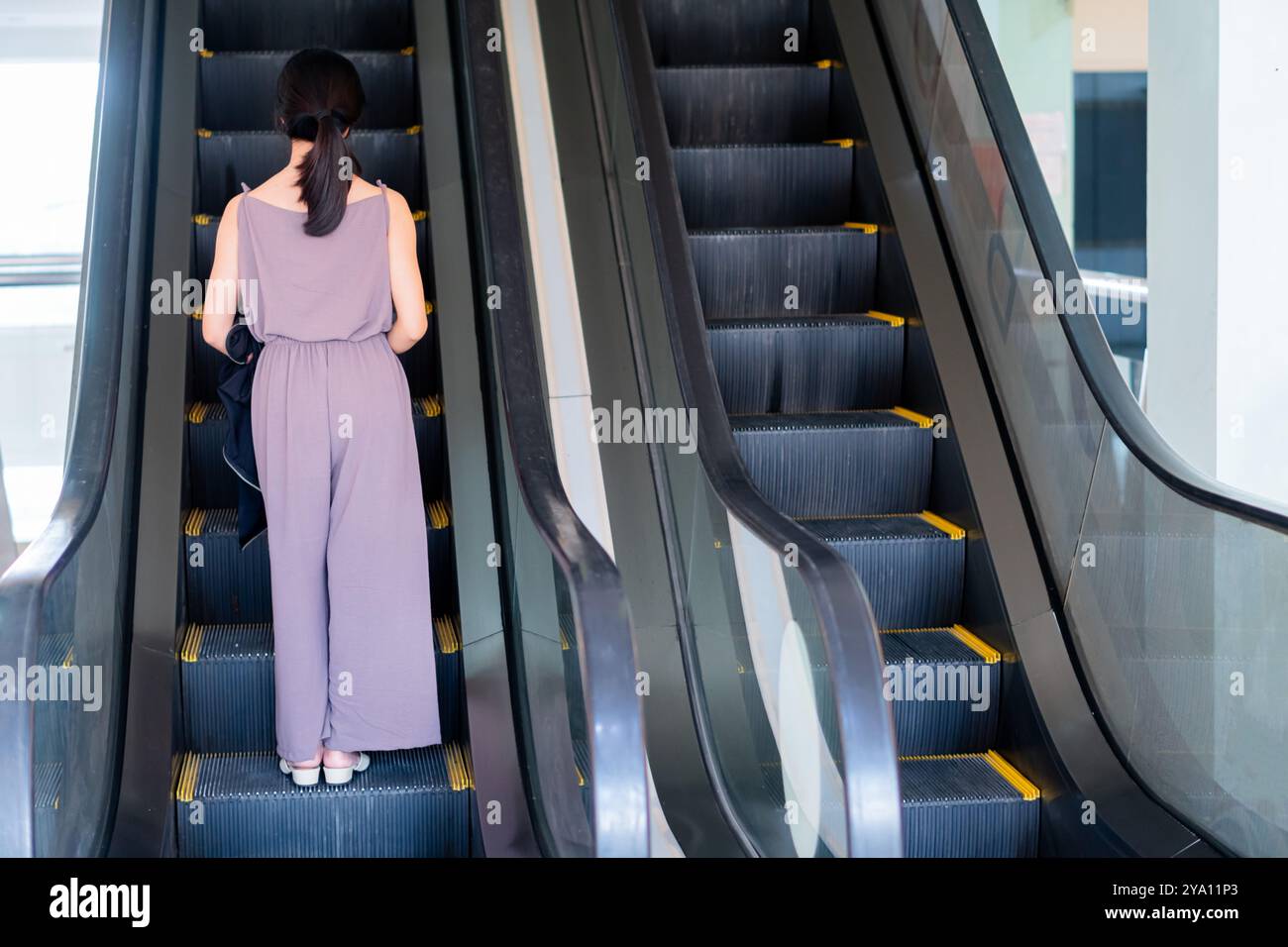 A woman in a gray jumpsuit is ascending an escalator. The escalator has ...