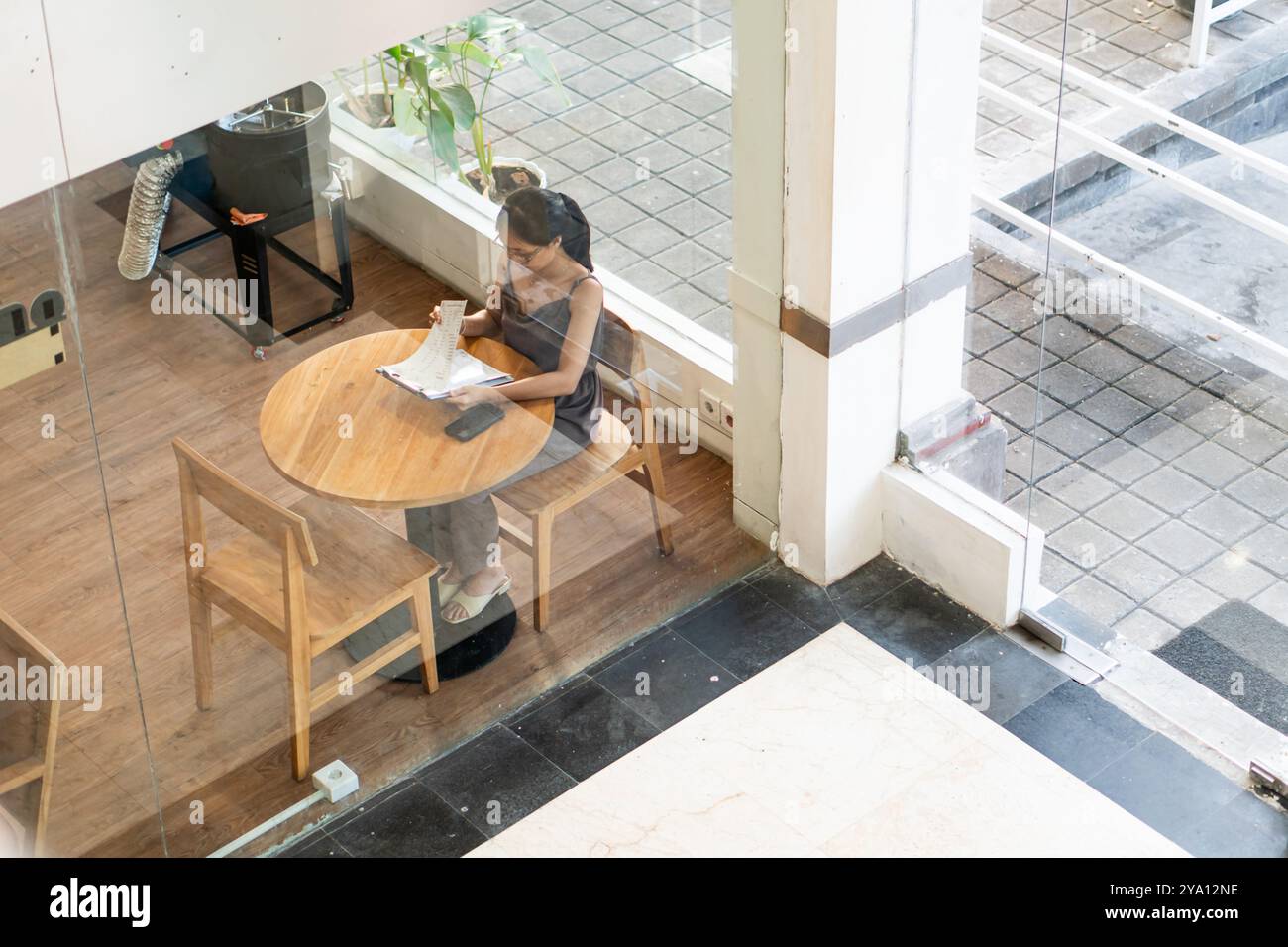 A woman sitting alone at a round wooden table in a modern cafe, reading ...