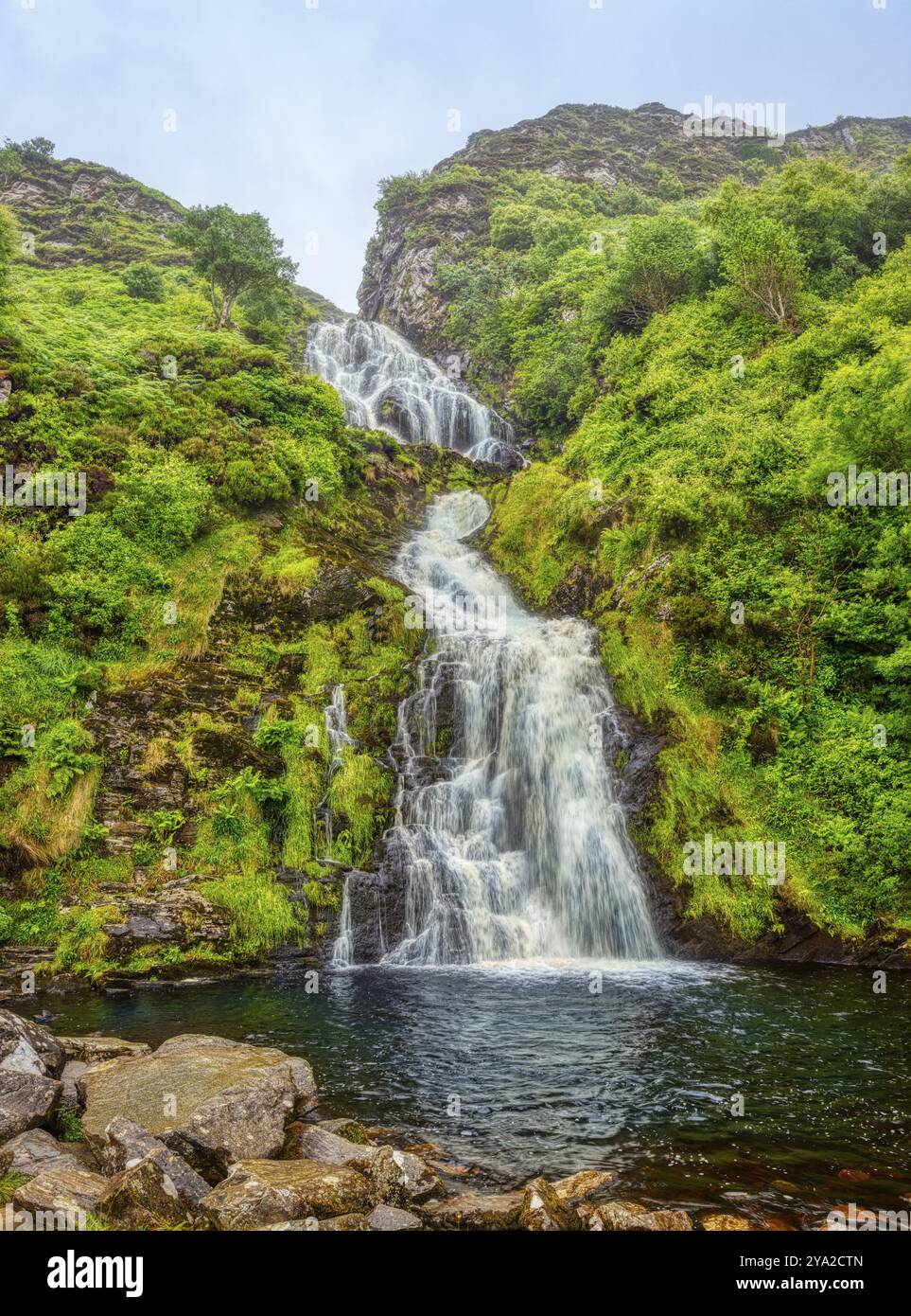 An impressive waterfall flows over rocks into a clear river surrounded ...