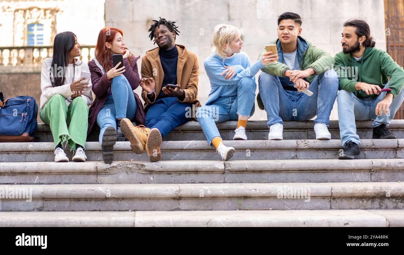 Group of diverse young students sitting on university steps, using smartphones and socializing ...