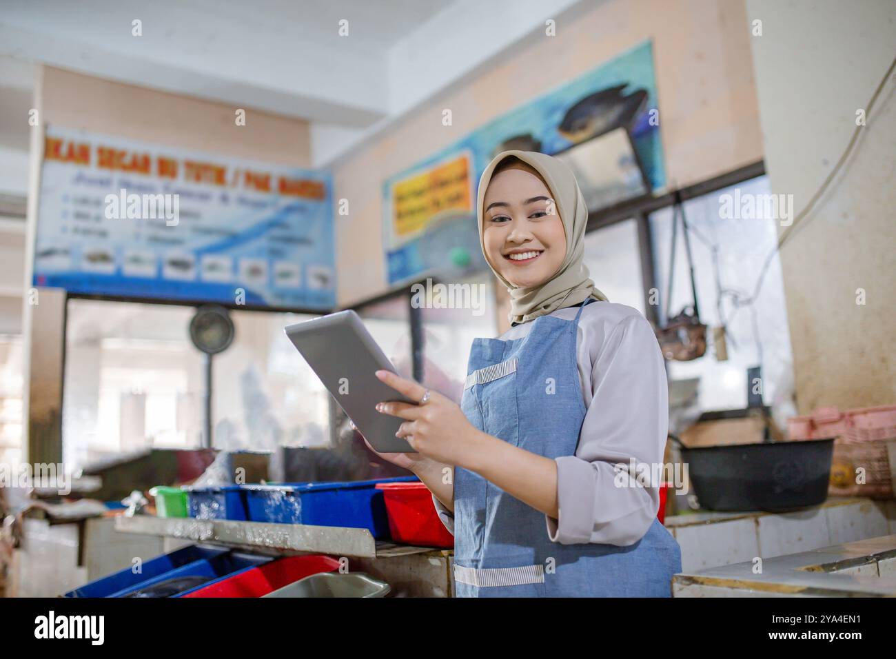 A Young Woman Dressed in a Hijab is Using a Tablet at a Bustling Fish ...