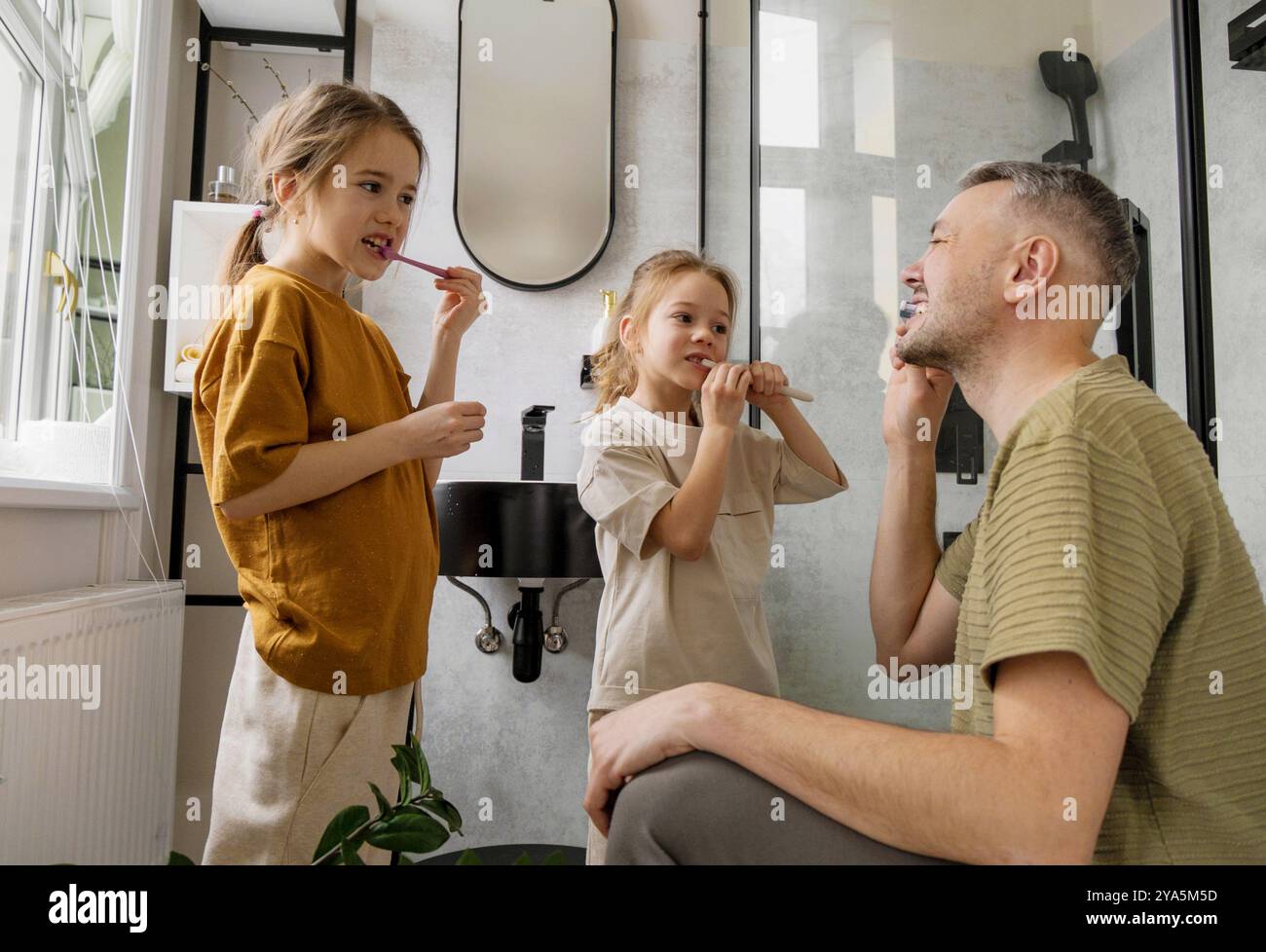A father teaches his two daughters how to brush their teeth in the ...