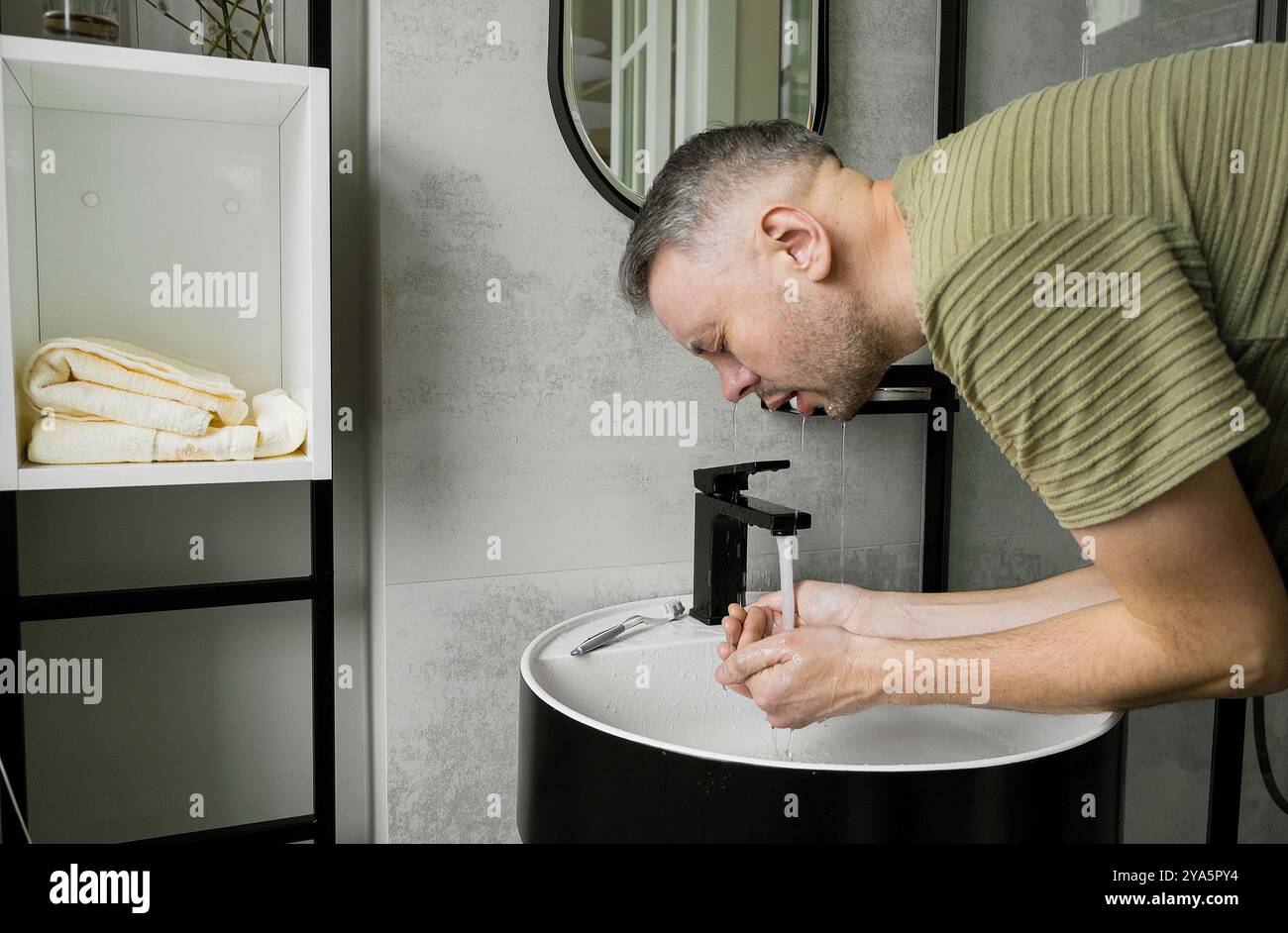 A man is washing his face in a modern bathroom. He is bending over the sink, with water running ...