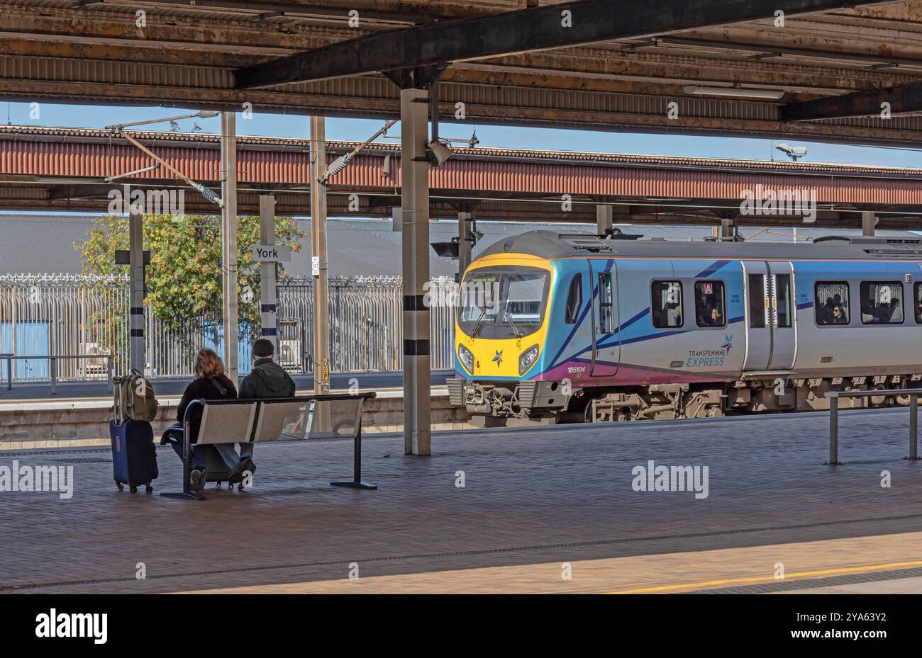 Two passengers sit on a bench in the shade and a train stands at a far ...