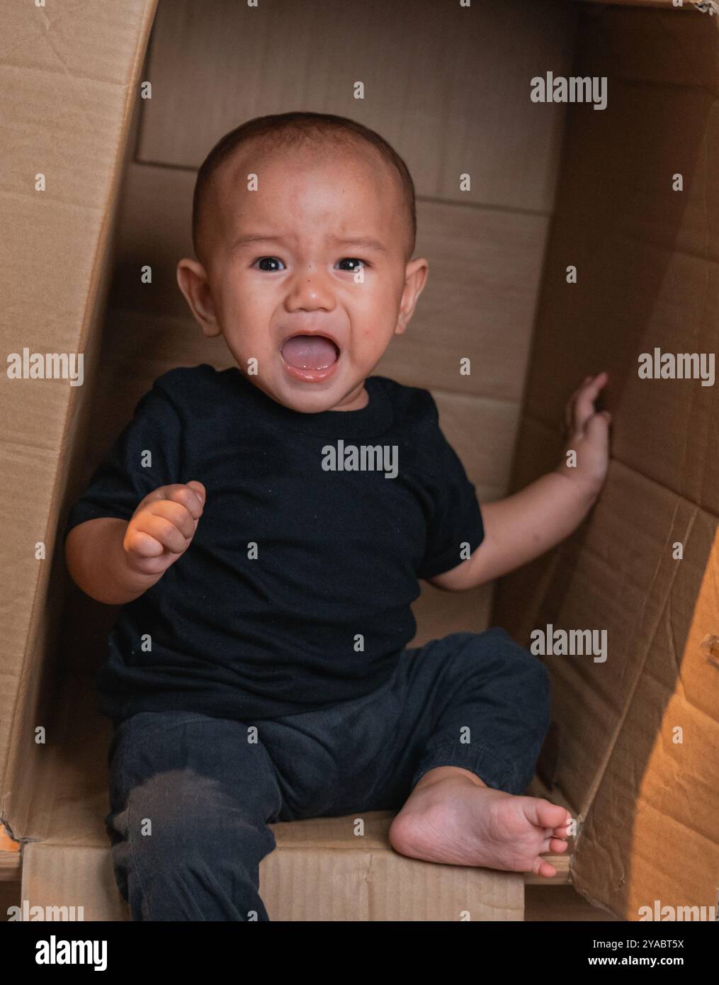A crying baby in a cardboard box shows a playful expression, capturing ...