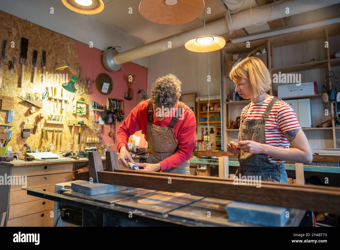 Craftsman demonstrate wooden skills to assistant in workshop. Master work on bed frame, apply ...
