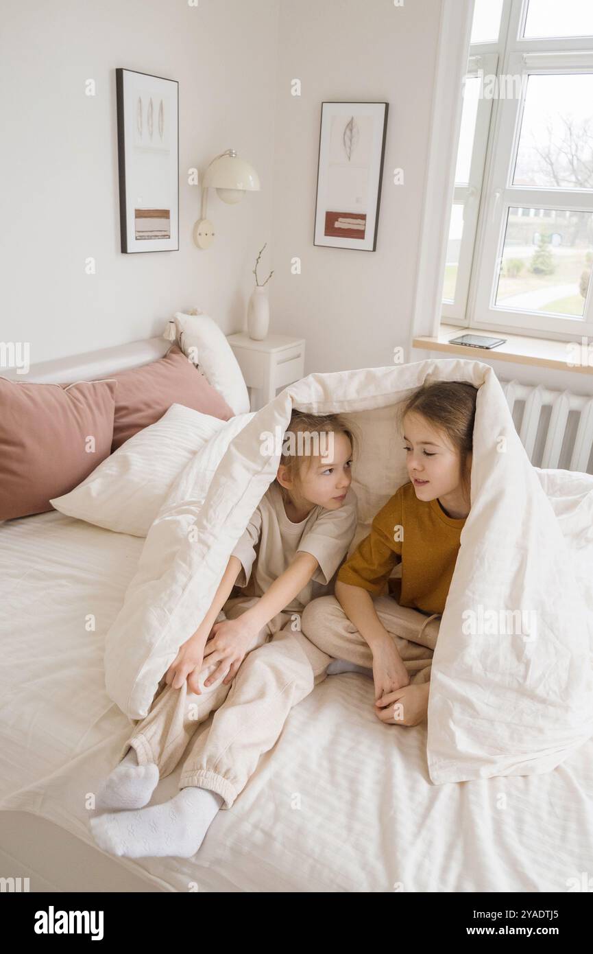 Two young girls, huddled under a blanket on a bed, share a quiet moment ...