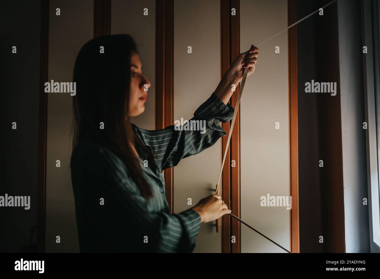 Woman adjusting window blinds in a dimly lit room with care Stock Photo ...