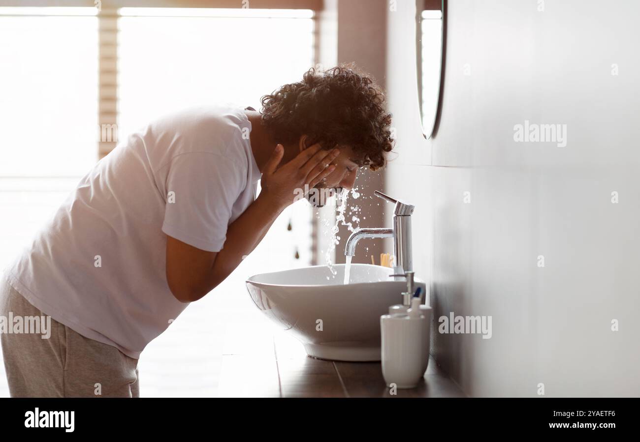Young indian man washing face with clear water from tap bending over ...