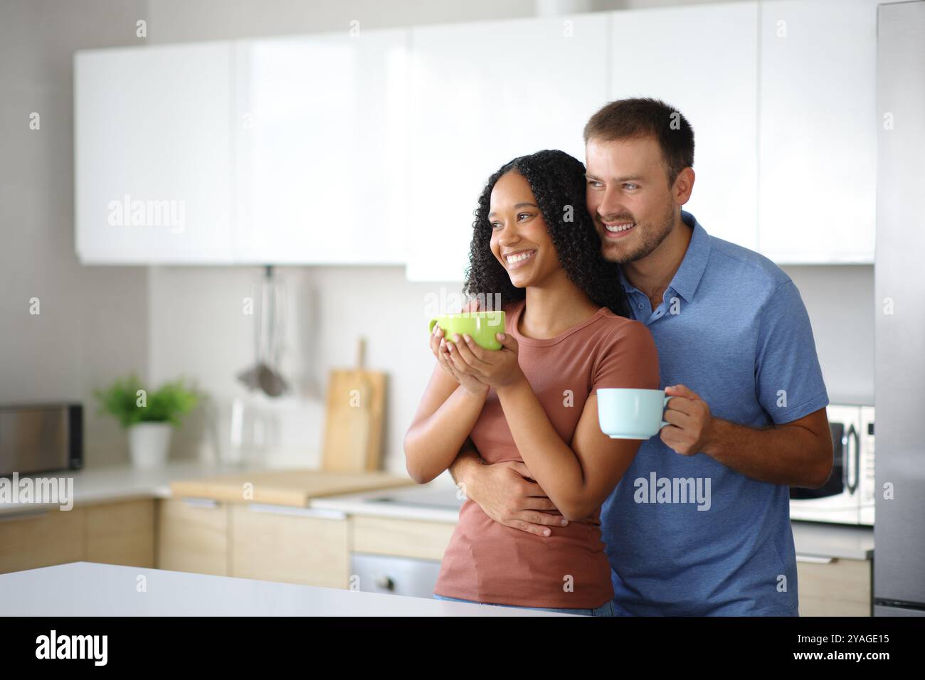 Happy interracial couple standing in the kitchen hugging and drinking ...