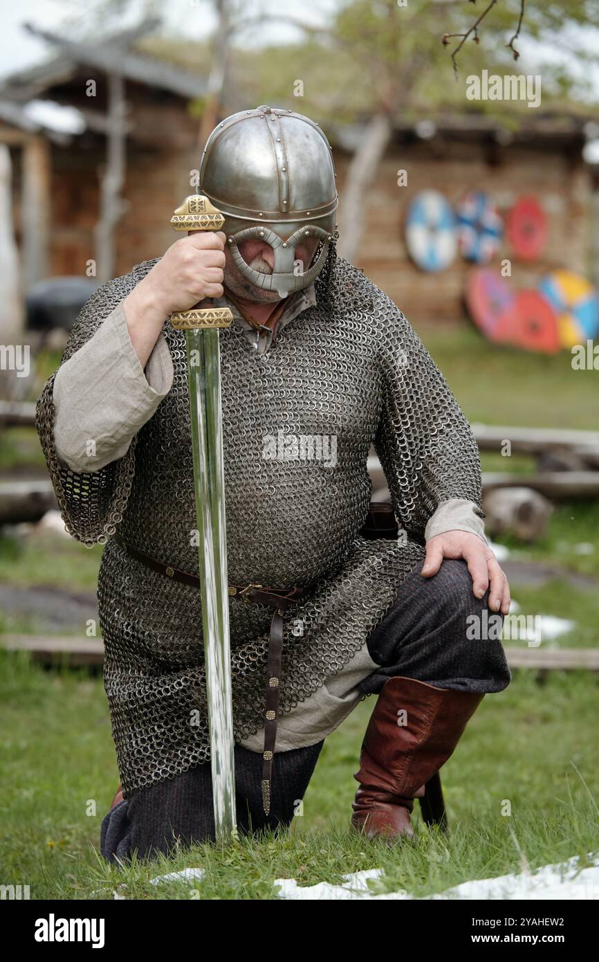 Medieval warrior dressed in chainmail and helmet kneeling on grass holding large sword in an ...