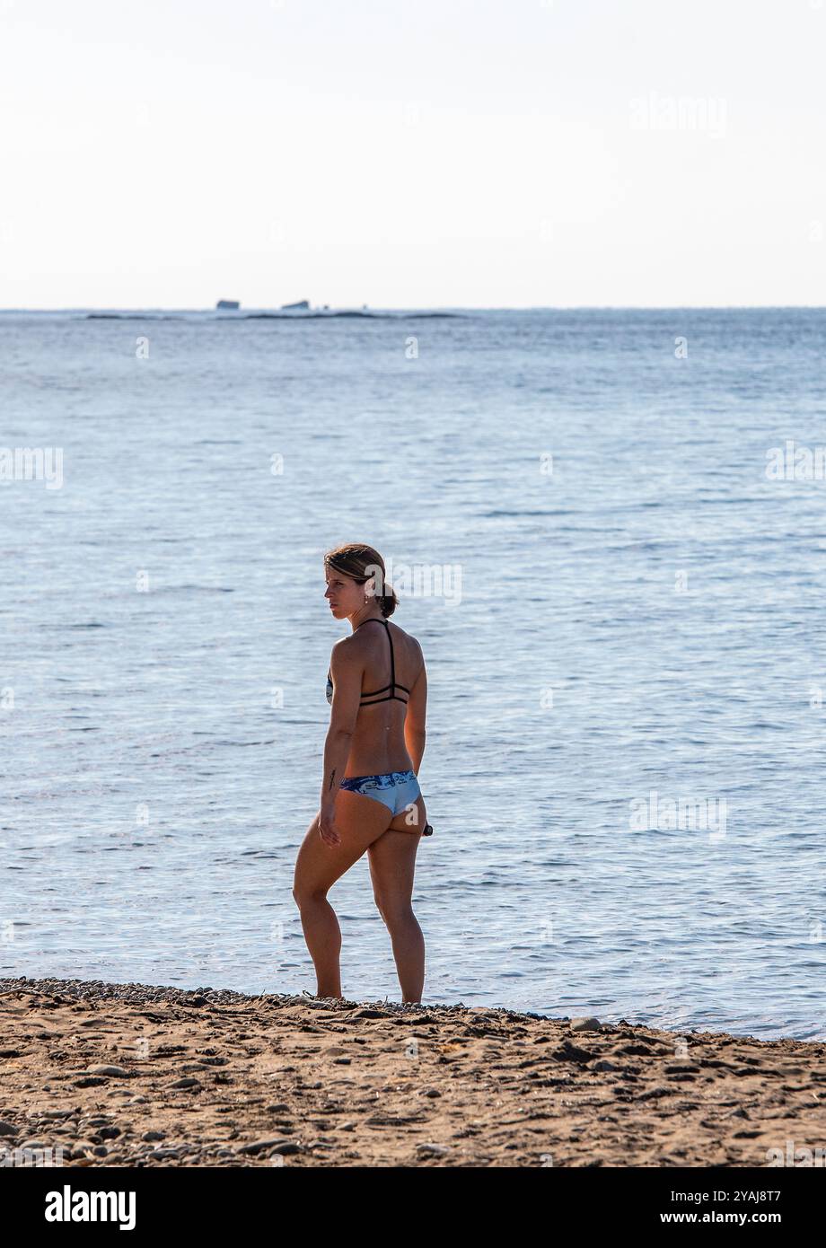 Attractive Slim Young Woman Walking Alone Along A Mediterranean Greek Beach Wearing A Blue