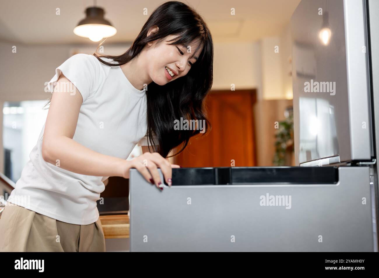 A charming, happy young Asian woman is open a fridge, looking for food ...