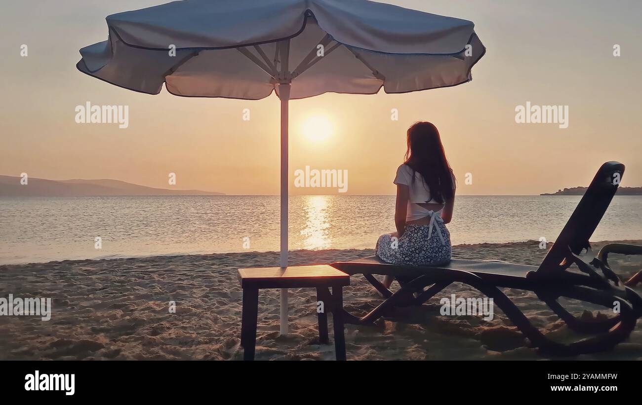 Aesthetic young woman seated on a sunbed at the beach watching the dawn ...