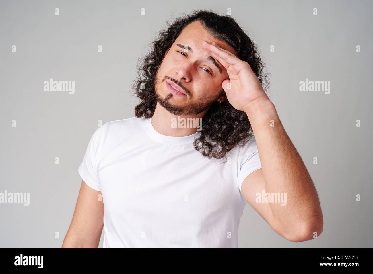 A young man with curly hair rubs his forehead in a casual indoor setting while expressing ...