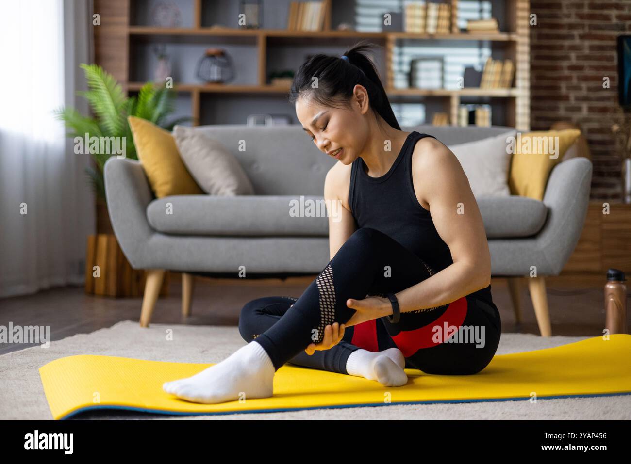 Asian woman injured leg during workout at home, seated on yoga mat ...